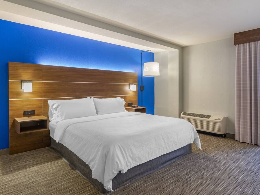Quarto de hotel com uma cama grande, uma mesa de cabeceira, uma luminária na parede e uma janela com cortinas.