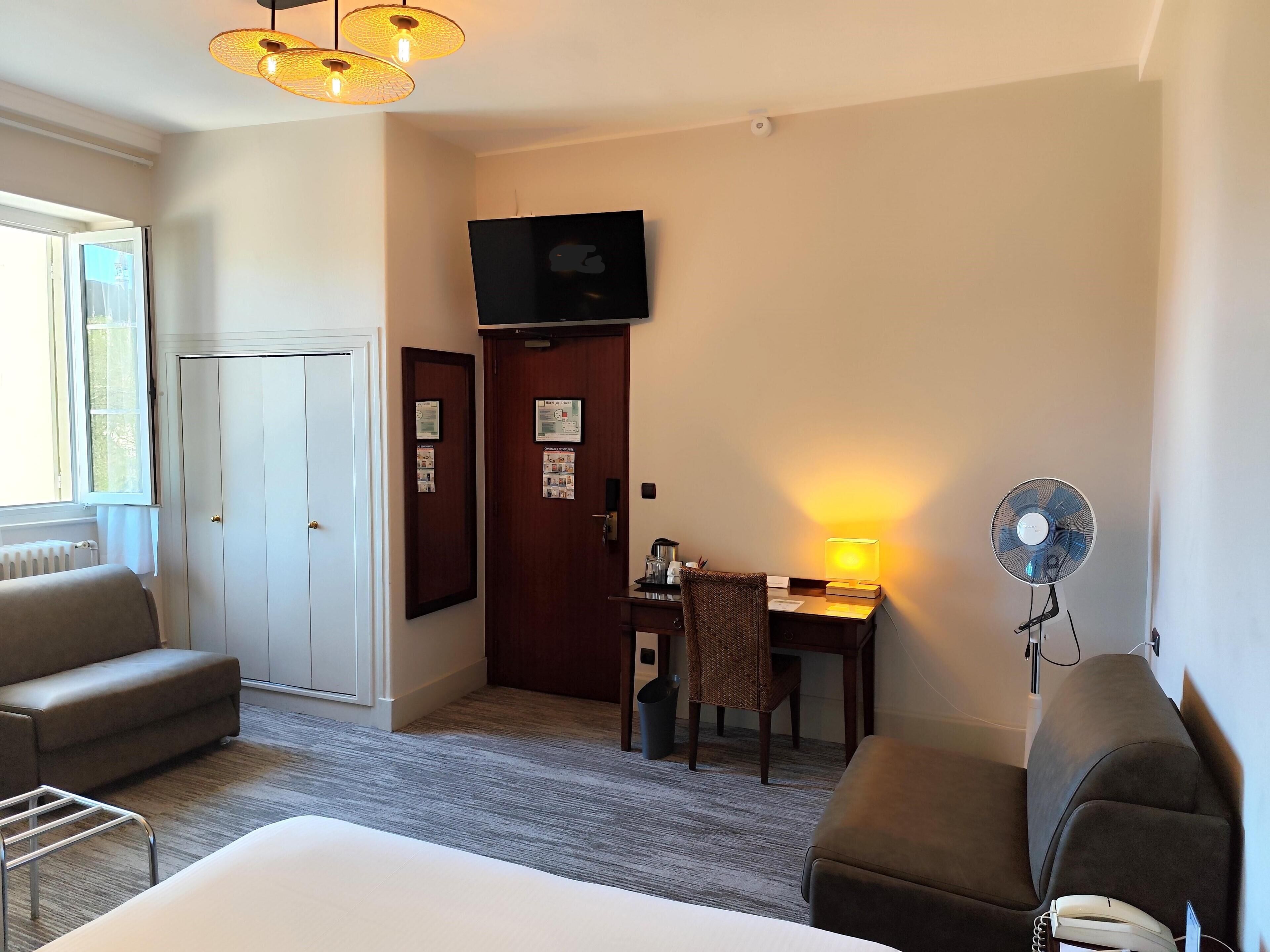 Quarto de hotel com uma cama, duas poltronas, uma escrivaninha com abajur, televisão e ventilador.