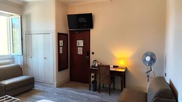 Quarto de hotel com uma cama, duas poltronas, uma escrivaninha com abajur, televisão e ventilador.