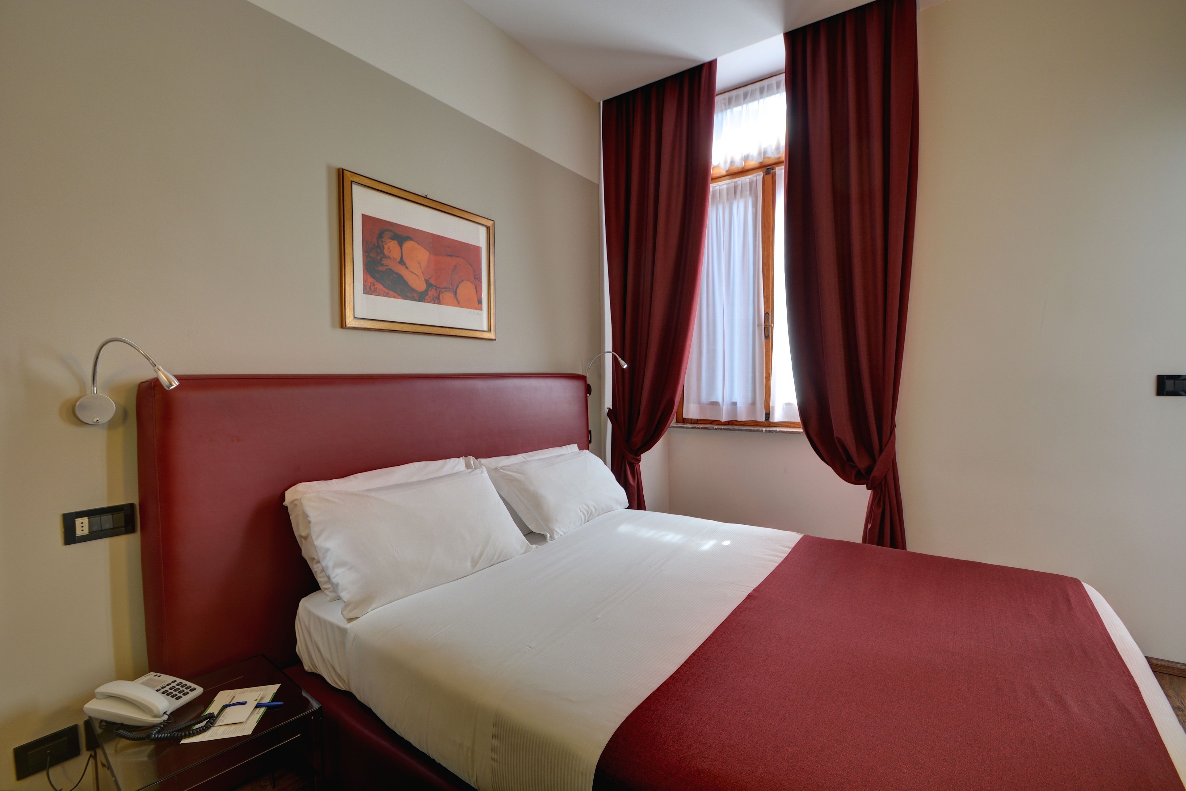 Quarto de hotel com uma cama grande, telefone de apoio, quadro na parede e janela com cortinas vermelhas.
