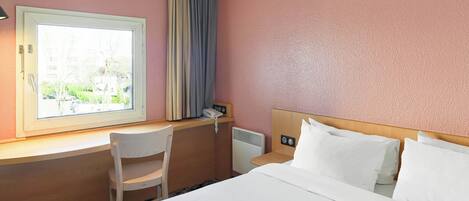Quarto de hotel com cama, escrivaninha com cadeira, janela com cortinas e aquecedor fixado na parede.