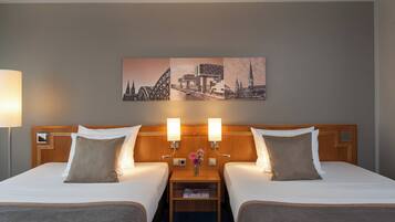 Quarto de hotel com duas camas, uma mesa de cabeceira, uma luminária e uma obra de arte na parede retratando paisagens urbanas.