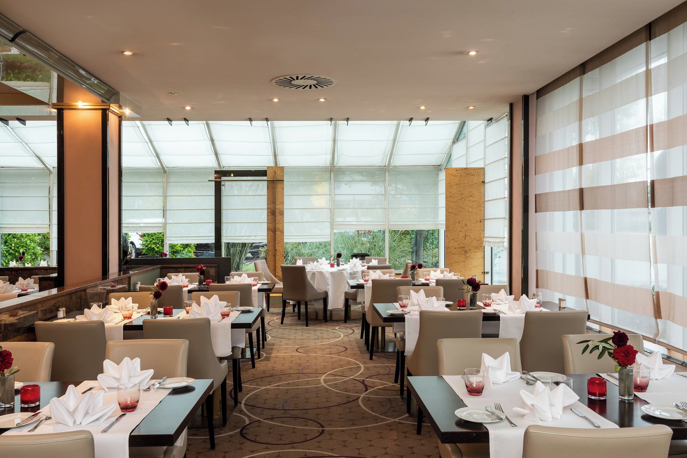 Restaurante com mesas preparadas para refeições, grandes janelas e vista para o verde.