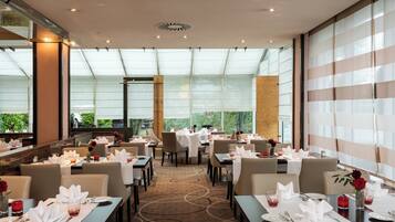 Restaurante com mesas preparadas para refeições, grandes janelas e vista para o verde.