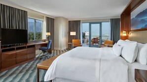 Quarto de hotel com uma cama grande, uma mesa com TV, uma cadeira, uma mesinha e vista para as montanhas pelas janelas.