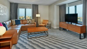 Quarto de hotel com uma janela grande, um sofá, uma mesa de centro, uma TV sobre um móvel de madeira e um tapete estampado.