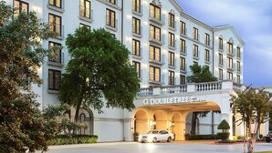 Um hotel grande e branco, com vários andares e varandas, apresenta uma entrada notável com toldo e um carro estacionado na frente.