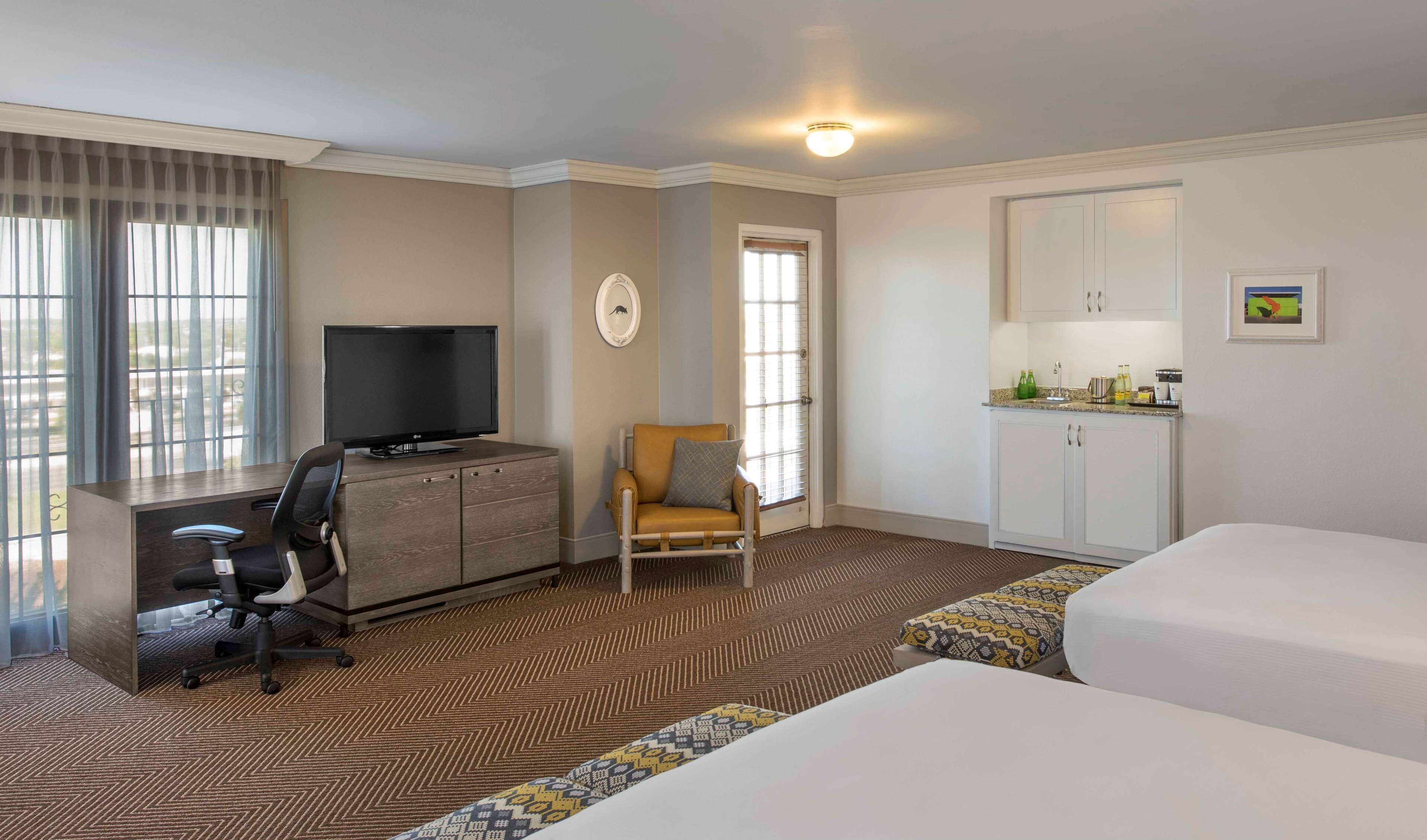 Quarto de hotel com televisão, escrivaninha, cadeira, cama, pequeno espaço de cozinha e janela com cortinas.