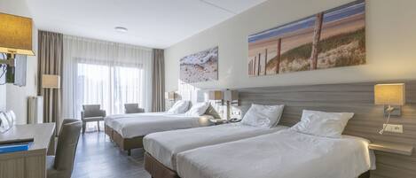 Quarto de hotel com duas camas, uma escrivaninha e uma cadeira. Há quadros nas paredes e uma janela com cortinas.