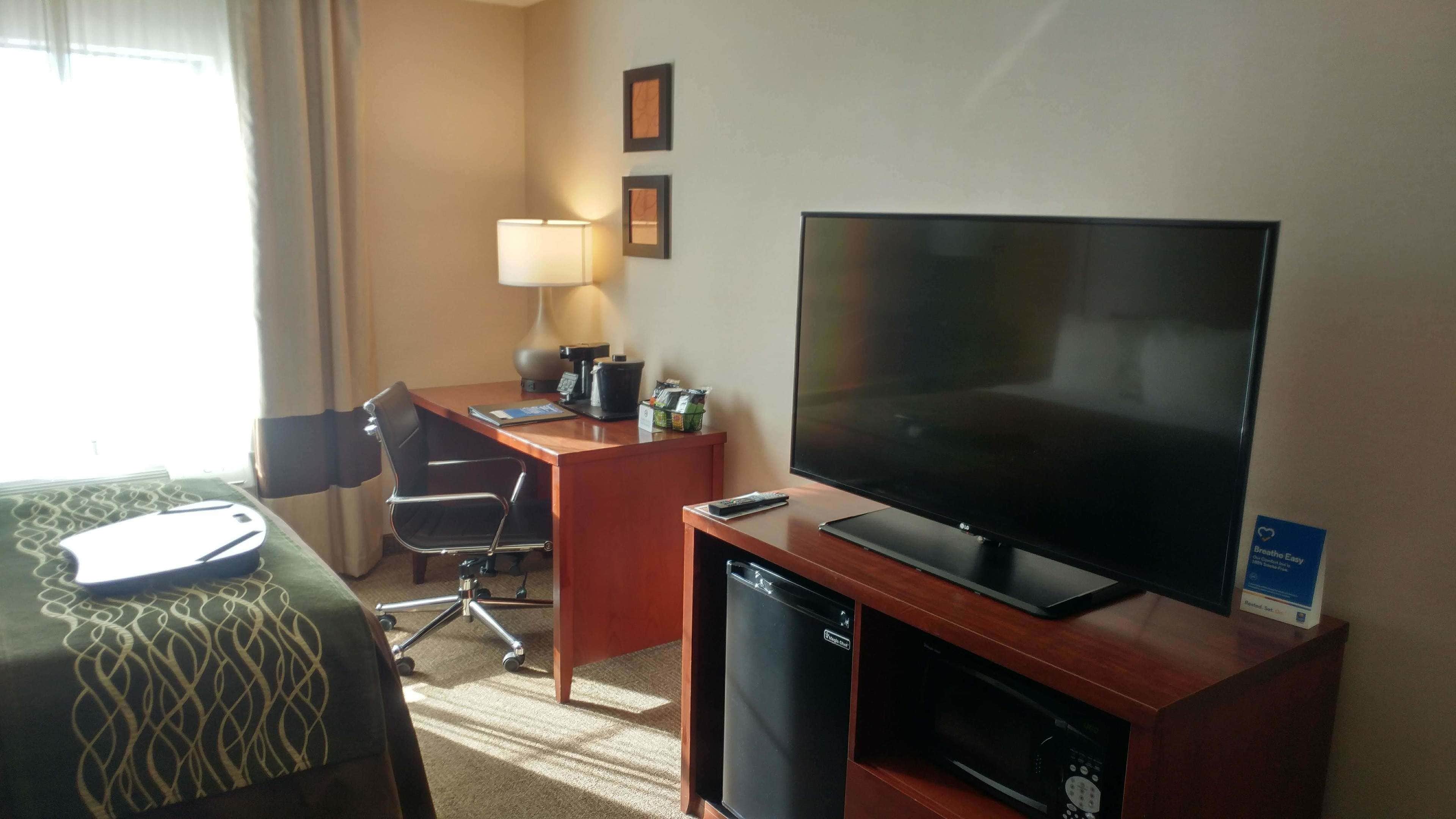Quarto de hotel com uma cama, uma escrivaninha com televisão, um abajur e um micro-ondas.