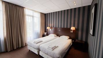 Quarto de hotel com duas camas, uma janela grande com cortinas, parede listrada e televisão fixada na parede.