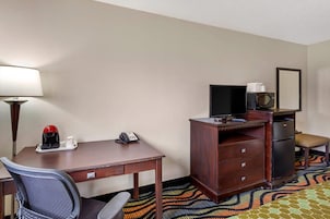 Quarto de hotel com escrivaninha, cadeira, luminária, telefone, televisão, micro-ondas, geladeira e tapete estampado.