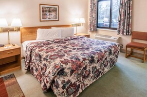 Quarto de hotel com uma cama grande, duas luminárias de cabeceira, uma mesinha de cabeceira, uma cadeira, uma janela com cortinas e um radiador.