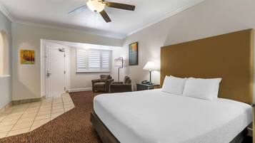 Quarto de hotel com uma cama grande, um ventilador de teto, uma escrivaninha com luminária, uma cadeira e um quadro na parede.