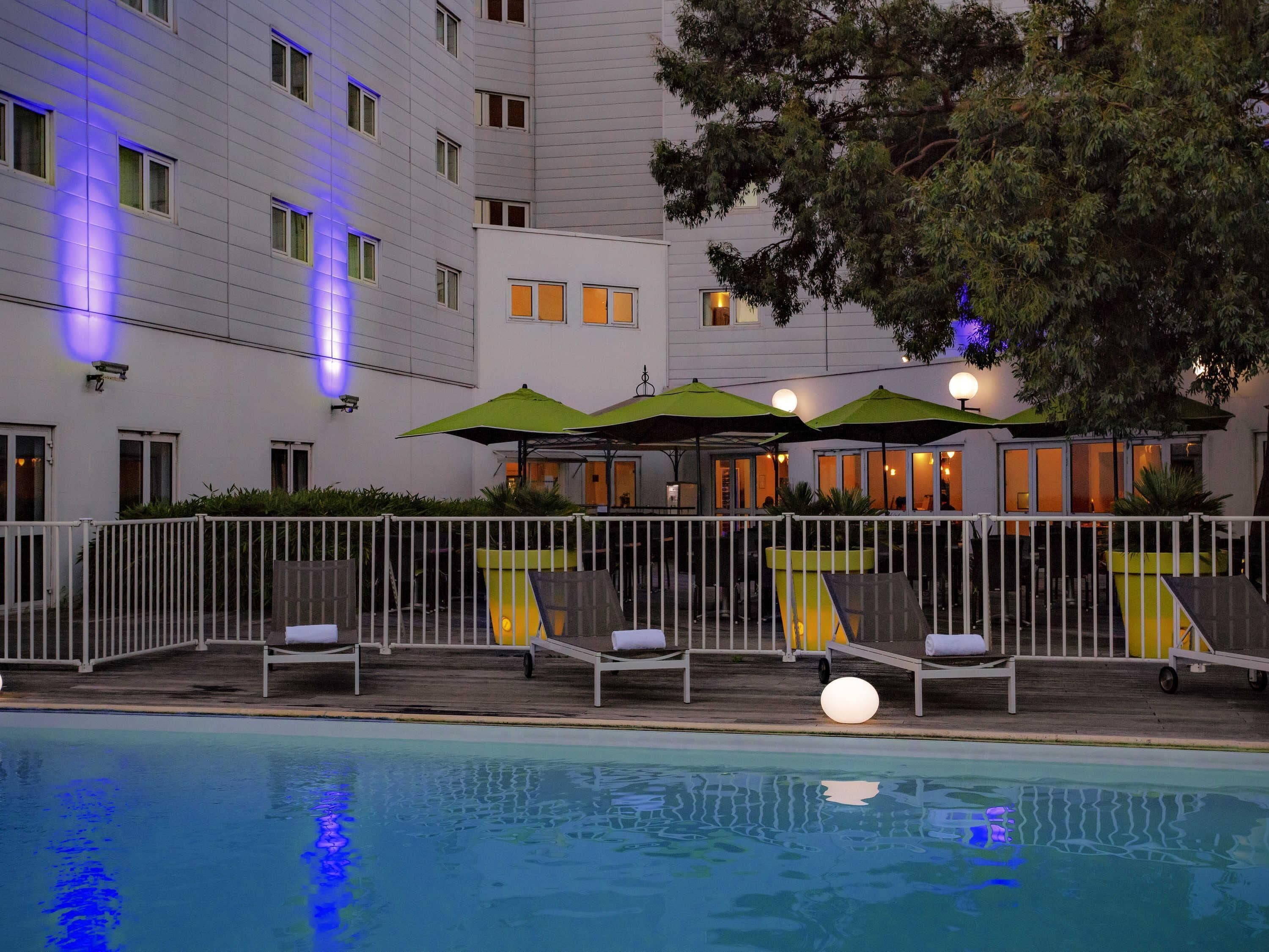 Uma área na beira da piscina com espreguiçadeiras, guarda-sóis e um edifício com várias janelas.