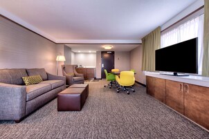 Quarto de hotel com um sofá cinzento, um banquinho castanho, uma televisão sobre um móvel de madeira e uma secretária com uma cadeira.