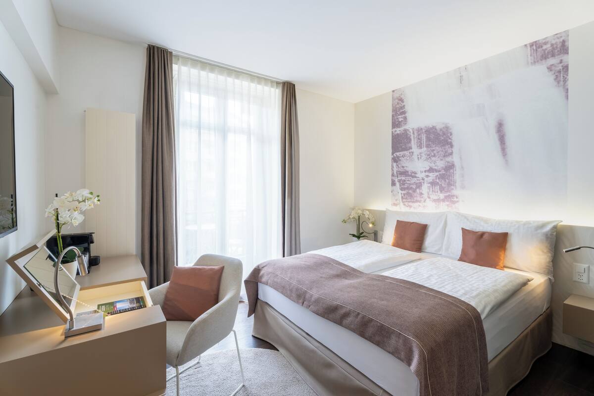 Quarto de hotel com uma cama, uma escrivaninha com cadeira, uma janela grande com cortinas e uma televisão presa à parede.
