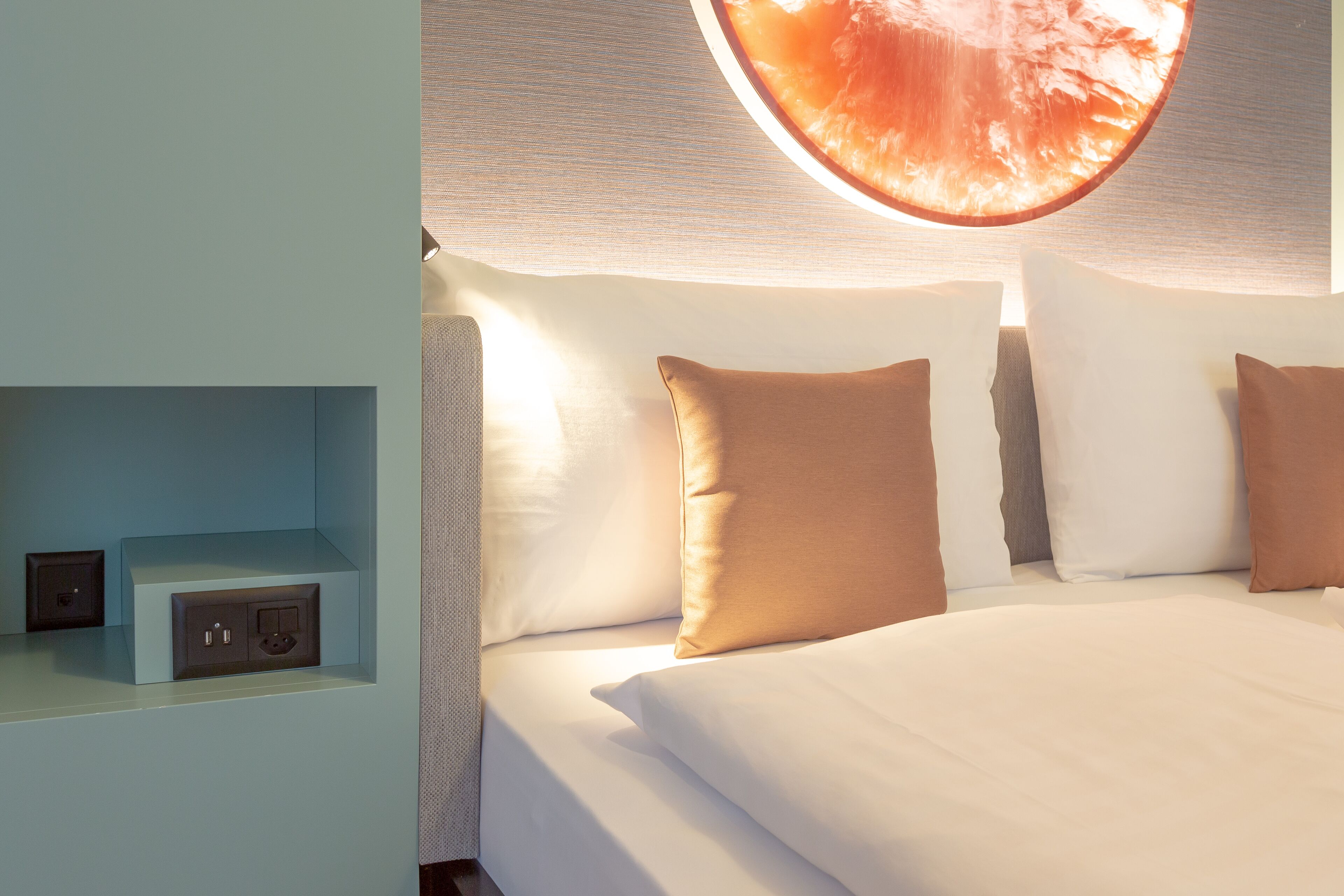 Quarto de hotel com cama, almofadas, mesa de cabeceira e luminária fixa na parede.