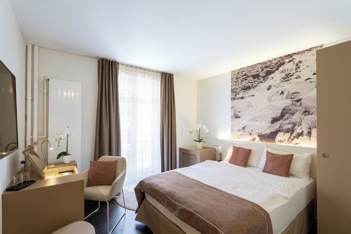 Quarto de hotel com uma cama grande, uma escrivaninha com cadeira, um espelho e uma janela com cortinas.