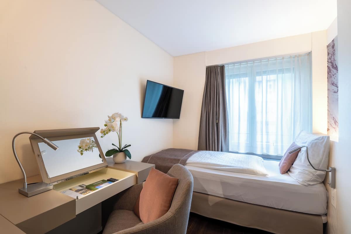 Quarto de hotel com escrivaninha, cadeira, cama, televisão e janela com cortinas.