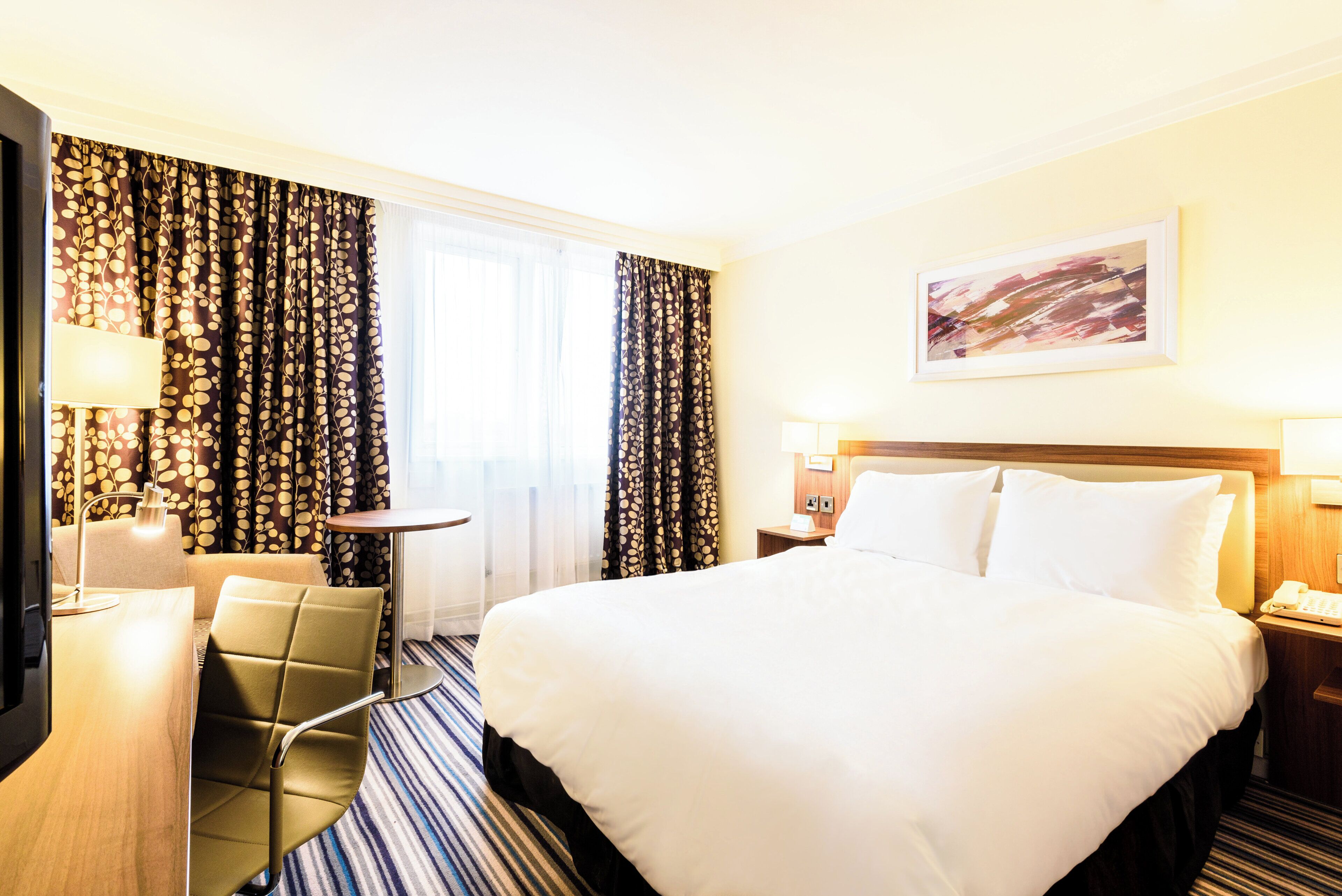 Quarto de hotel com uma cama, escrivaninha, cadeira, mesa e um quadro na parede.