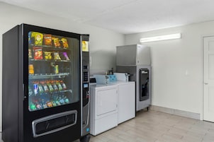 Máquina de vendas com lanches e bebidas, máquina de lavar roupa e secadora em um espaço com piso ladrilhado e porta branca.