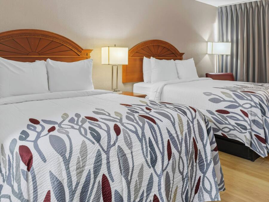 Quarto de hotel com duas camas, cabeceiras de madeira, roupa de cama branca e uma janela grande com cortinas.