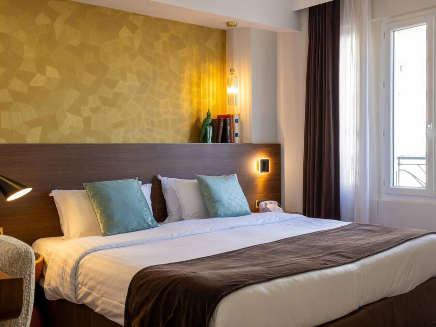 Quarto de hotel com uma cama grande, escrivaninha com abajur, telefone e janela com cortinas.