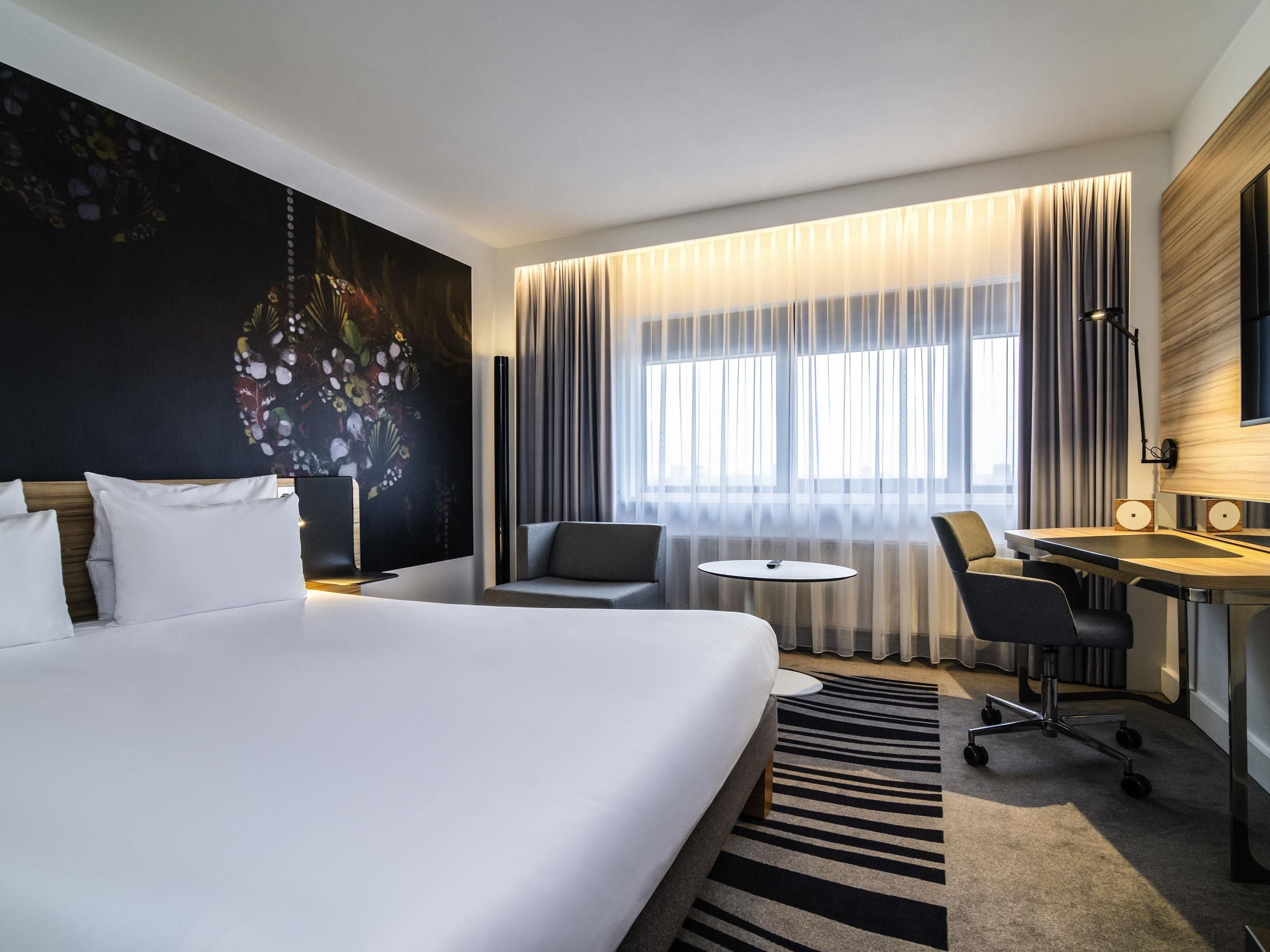Quarto de hotel com uma cama grande, uma escrivaninha com dois alto-falantes, uma cadeira, uma mesinha redonda e uma janela ampla com cortinas.