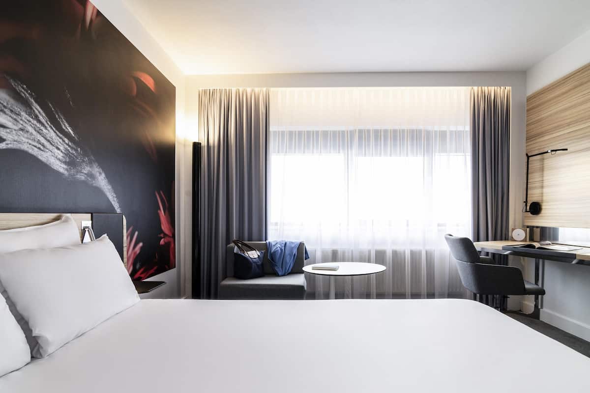 Quarto de hotel moderno com uma cama grande, uma escrivaninha com cadeira, uma mesinha redonda pequena e uma janela ampla com cortinas.