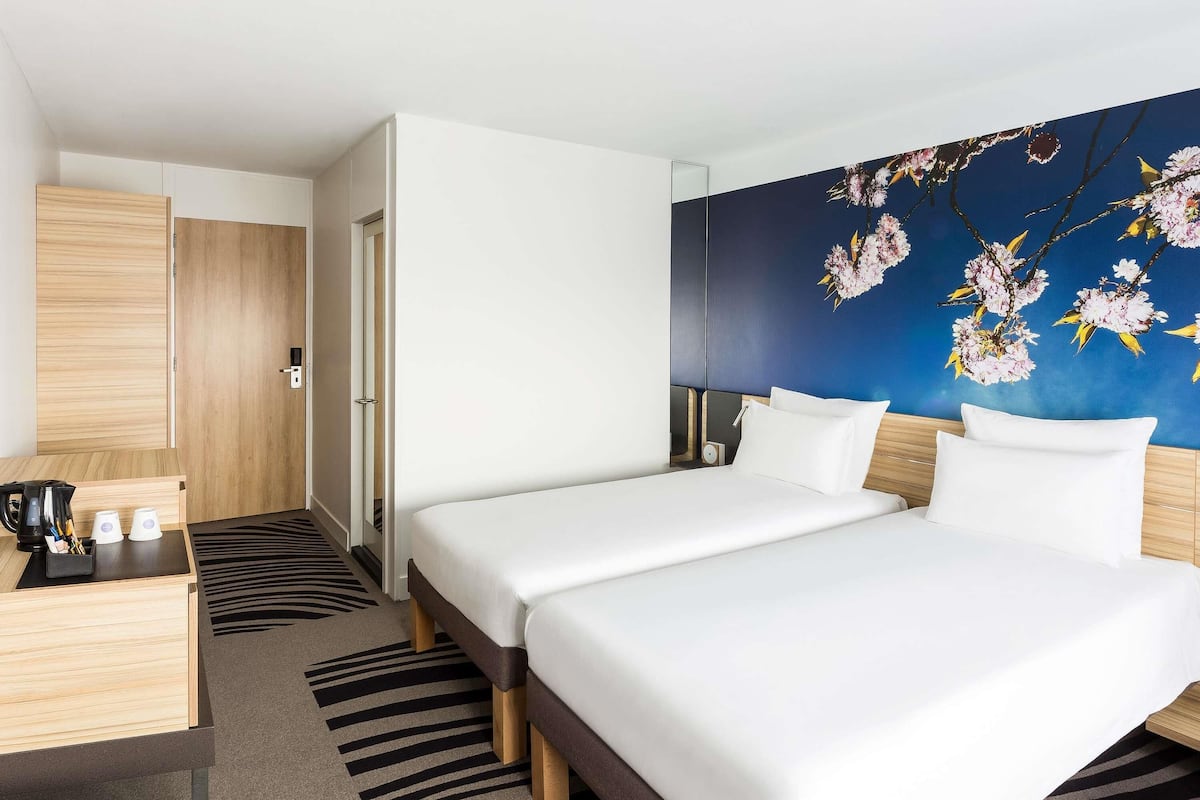 Quarto de hotel com duas camas, uma secretária de madeira, uma cafeteira e uma parede com um mural floral.