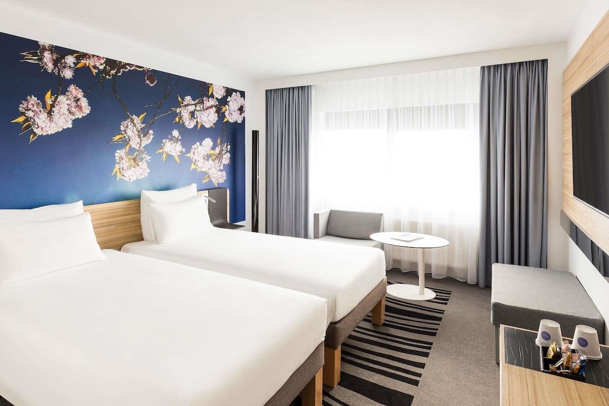 Quarto de hotel com duas camas, uma mesa redonda pequena, um sofá, uma televisão e uma janela com cortinas.