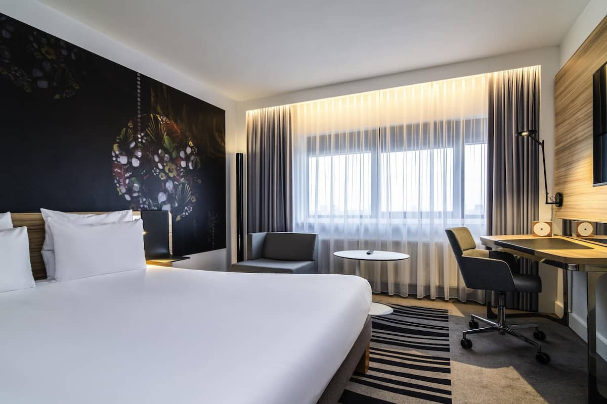 Quarto de hotel com uma cama grande, uma escrivaninha com dois alto-falantes, uma cadeira, uma mesinha redonda e uma janela ampla com cortinas.