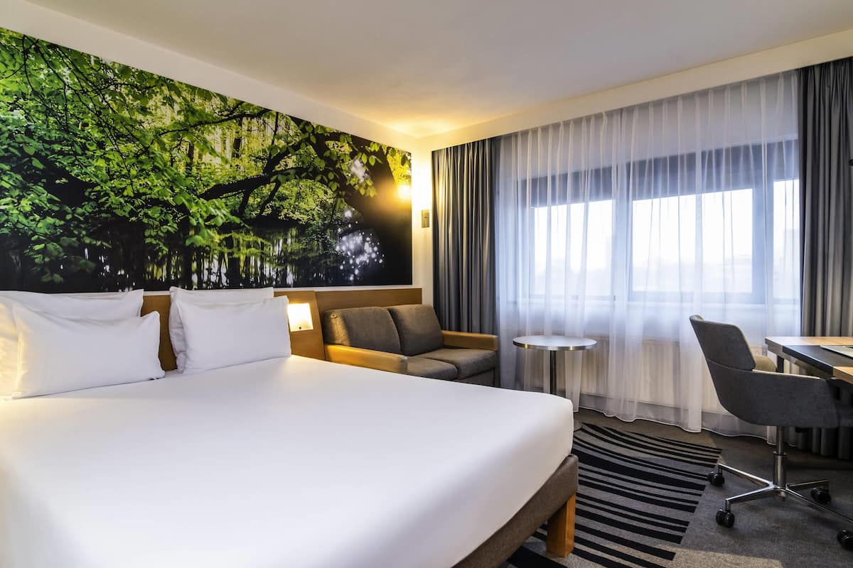 Quarto de hotel com uma cama grande, um sofá, uma escrivaninha e uma cadeira. Há uma janela com cortinas finas e vista para o exterior.