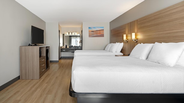 Quarto de hotel com duas camas, uma TV, um armário de madeira e um banheiro visível ao fundo.