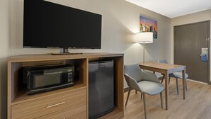 Quarto de hotel compacto com TV de tela plana, micro-ondas, mini-geladeira, mesa de jantar com duas cadeiras e um quadro na parede.