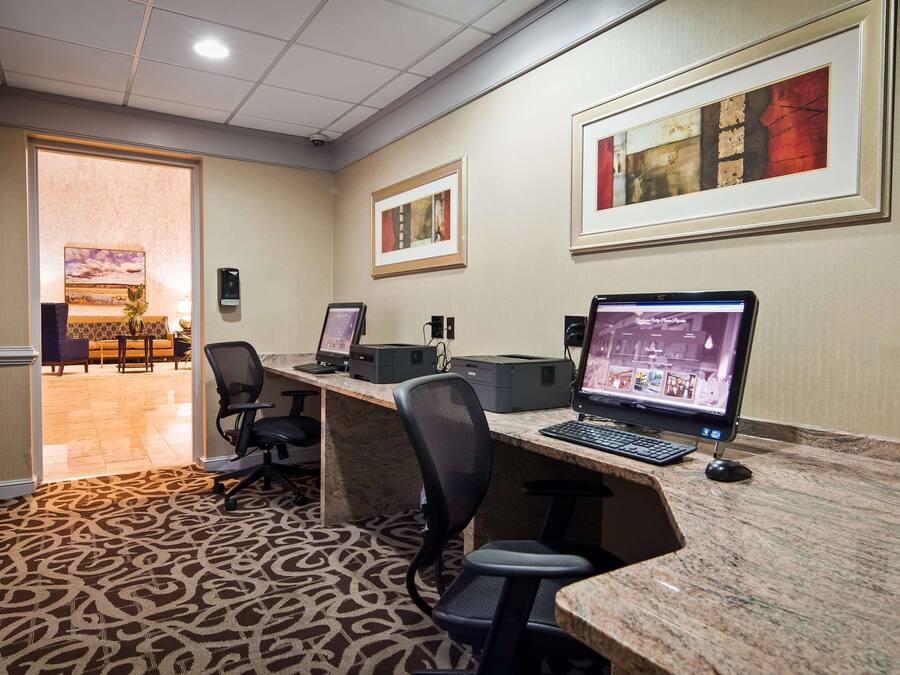 Quarto de hotel com escrivaninha, duas cadeiras, computador e impressora. Há um quadro na parede e uma porta que dá para um hall com sofá e uma pintura.