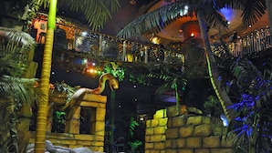 Um espaço interior com tema tropical, apresentando estruturas de pedra, palmeiras e uma piscina.
