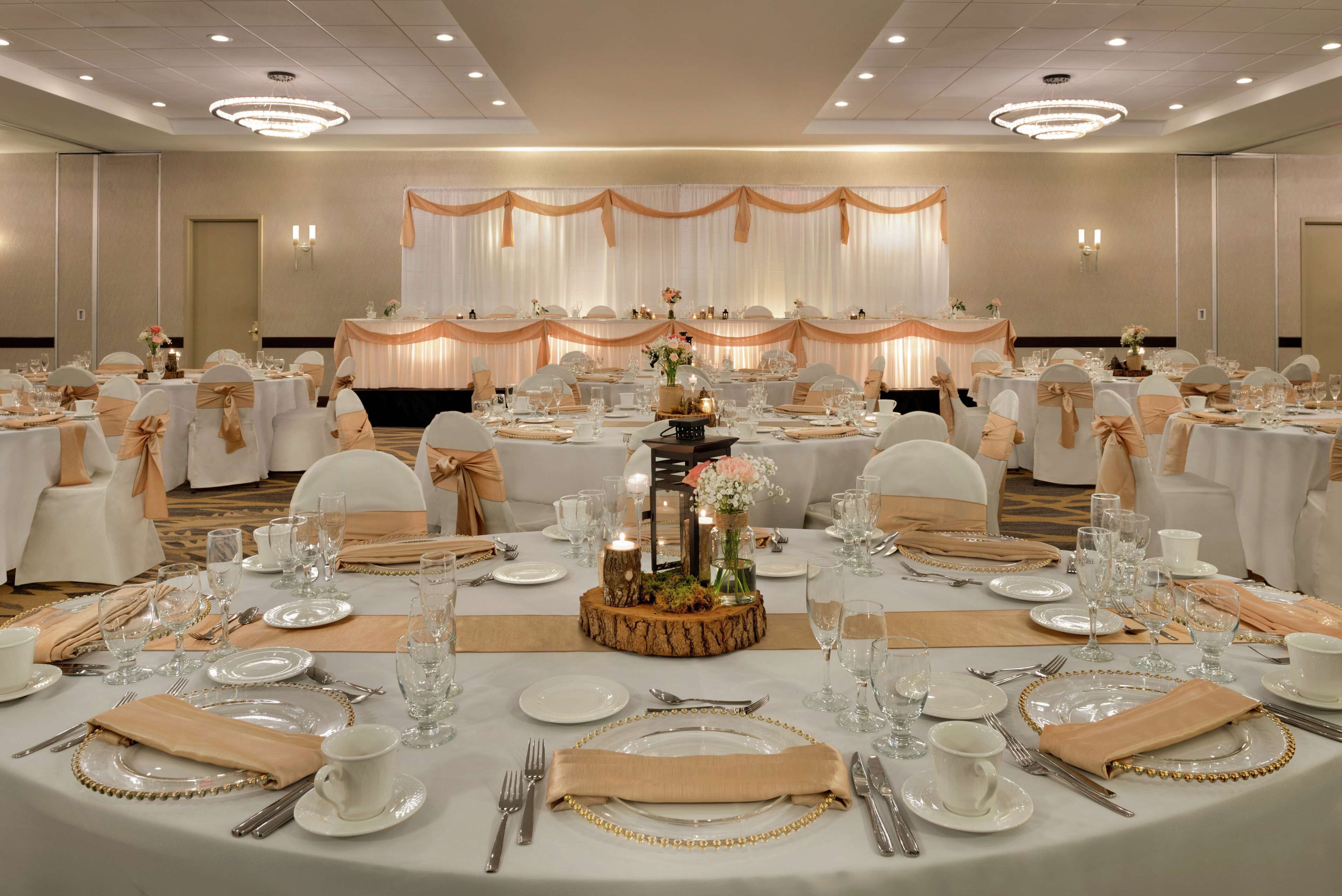 Salão de festas com mesas redondas dispostas para um evento formal, com toalhas brancas, detalhes dourados e arranjos florais.