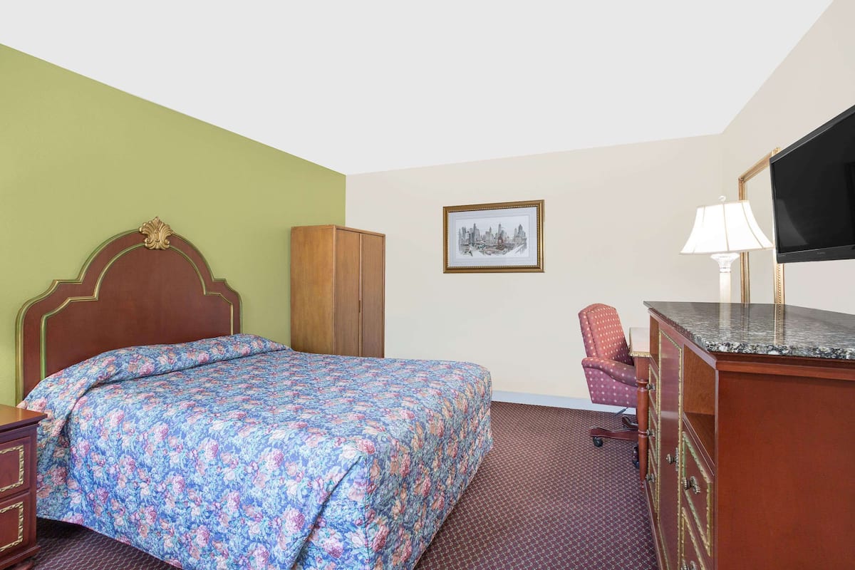 Quarto de hotel com cama, escrivaninha, cadeira, guarda-roupa, televisão e um quadro na parede.
