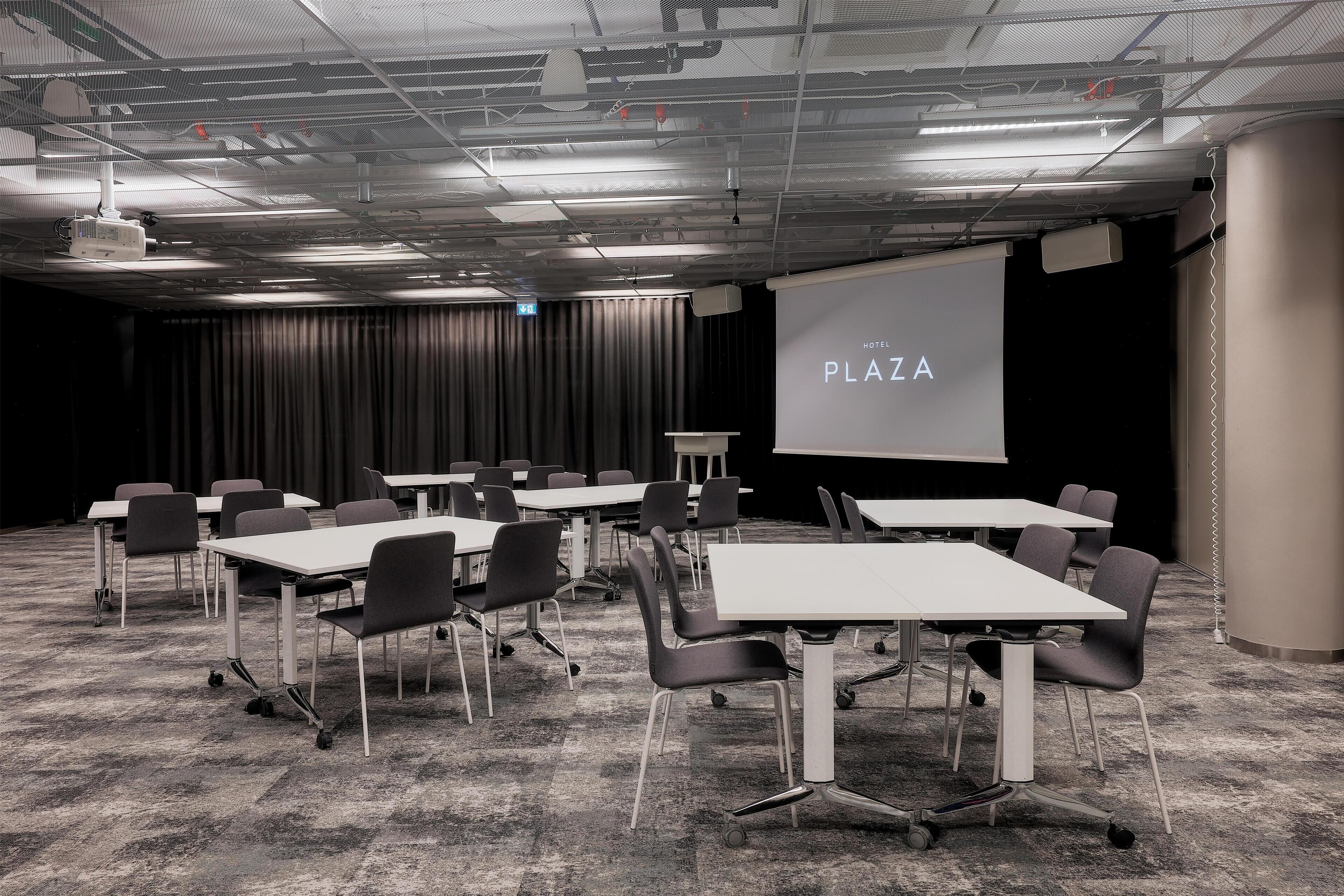 Uma sala de conferências com mesas, cadeiras, uma tela de projetor exibindo "PLAZA" e um fundo de cortina escura.