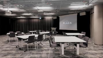 Uma sala de conferências com mesas, cadeiras, uma tela de projetor exibindo "PLAZA" e um fundo de cortina escura.
