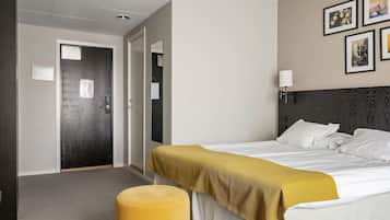 Quarto de hotel com cama, um banquinho amarelo, luminária fixada na parede e quadros na parede.