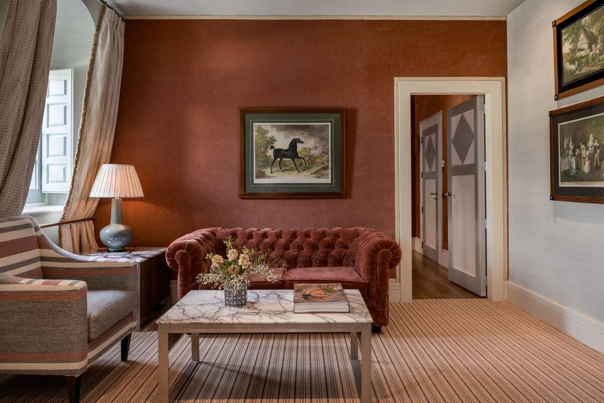 Um quarto com uma poltrona listrada, um sofá estofado, uma mesa de centro de mármore com um livro e flores, uma pintura de cavalos emoldurada e uma porta com padrões geométricos.