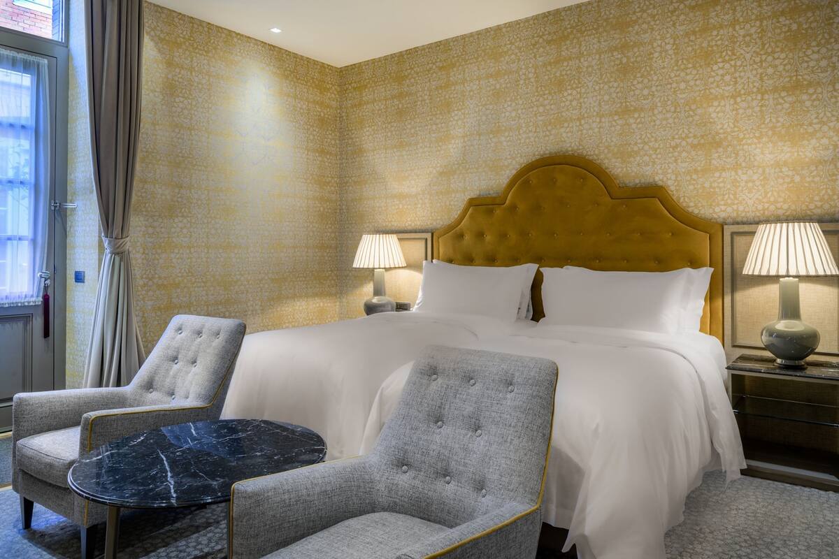 Quarto de hotel com uma cama grande, duas poltronas, uma mesa pequena e duas luminárias de cabeceira.