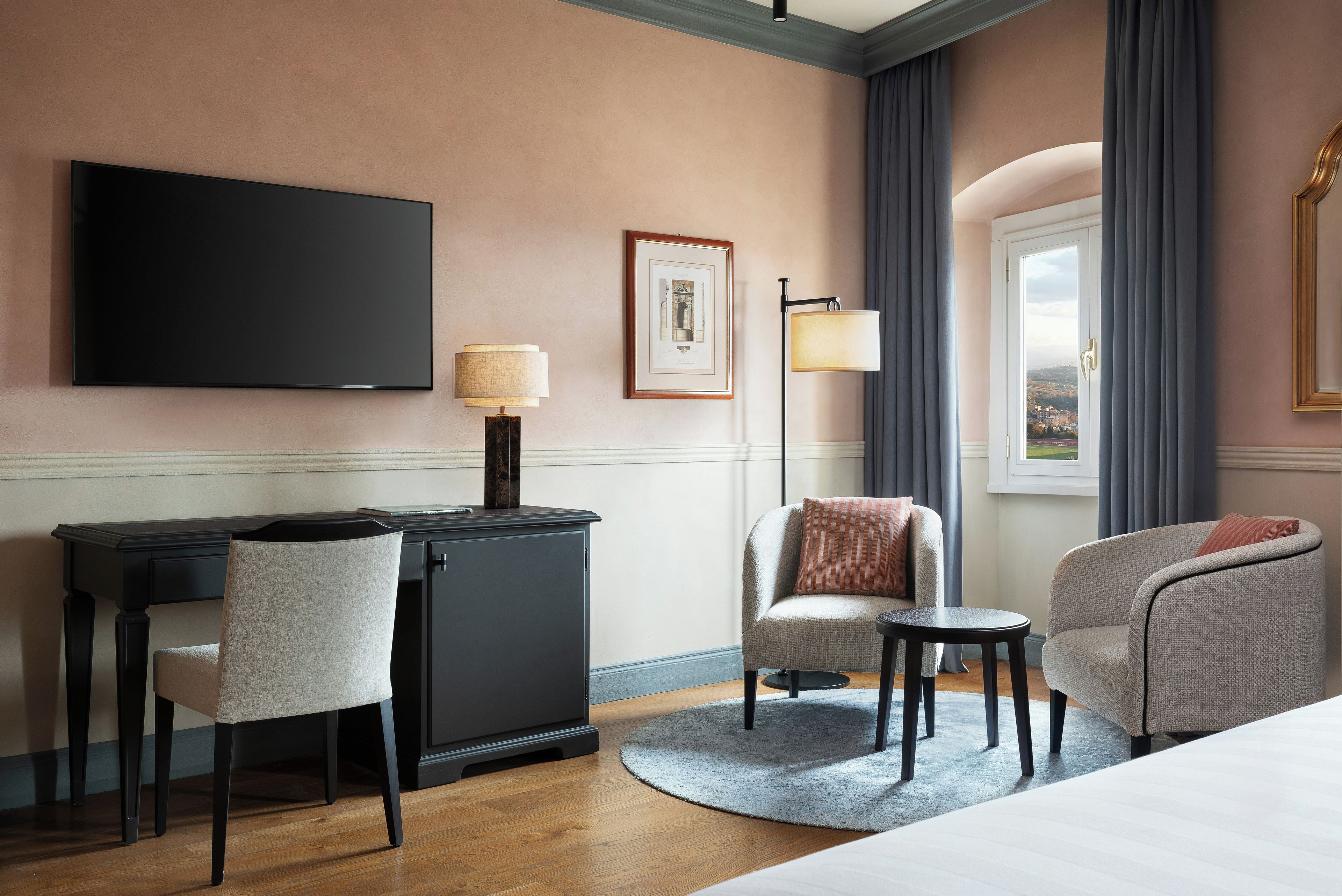 Quarto de hotel com televisão de ecrã plano, secretária com cadeira, sofá, mesa pequena e candeeiro.