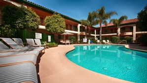 Área de piscina com espreguiçadeiras, piscina transparente e edifício com telhado de telhas vermelhas.