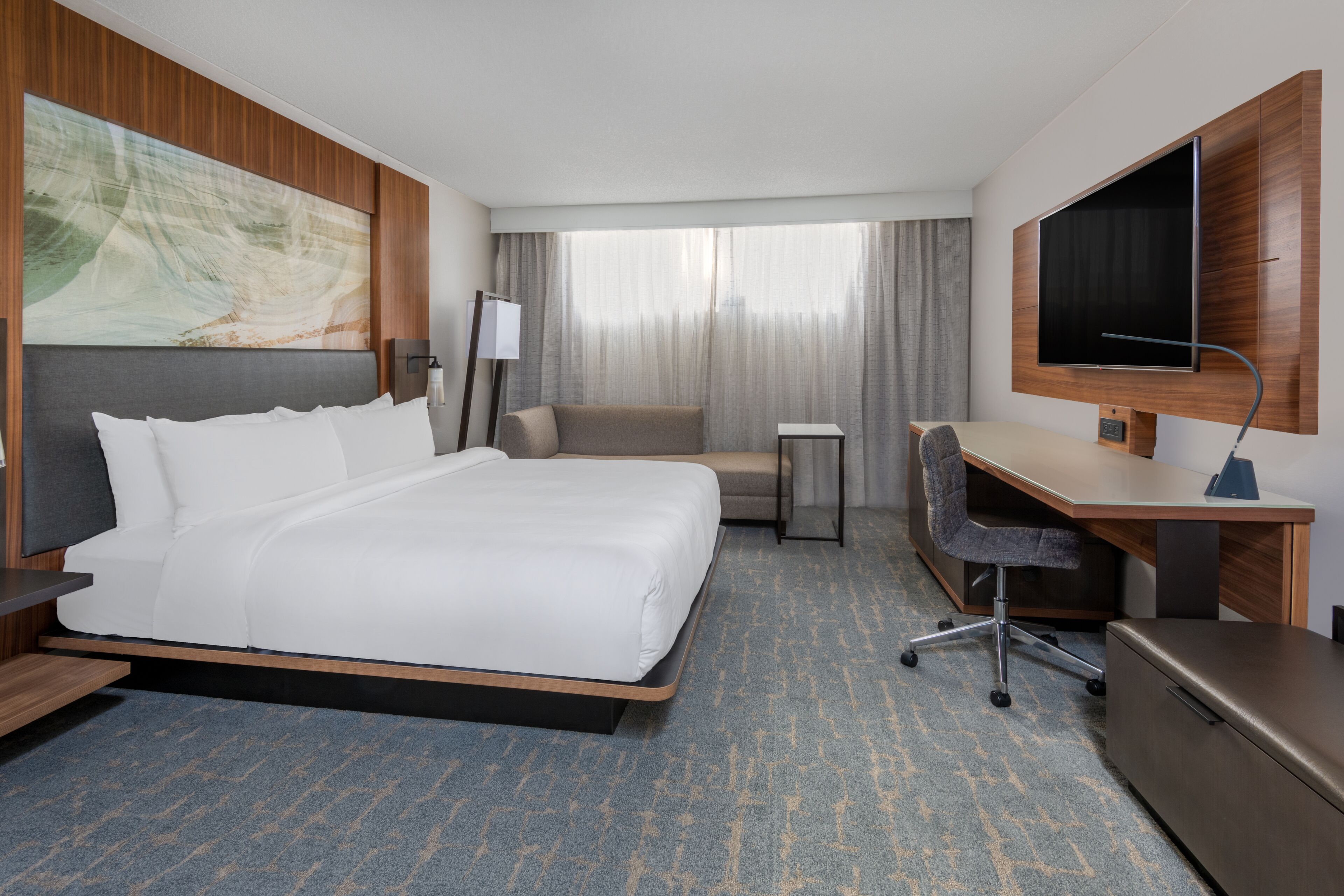 Quarto de hotel com uma cama grande, escrivaninha, cadeira, televisão e janela com cortinas.
