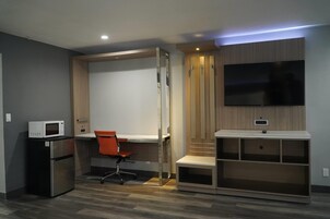 Quarto compacto com cozinha compacta, uma escrivaninha com cadeira laranja, TV fixada na parede e estantes embutidas.