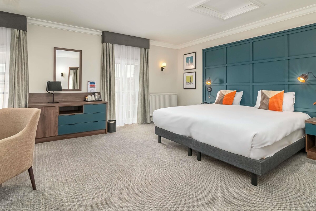 Quarto de hotel com cama, escrivaninha, cadeira e uma janela grande com cortinas.